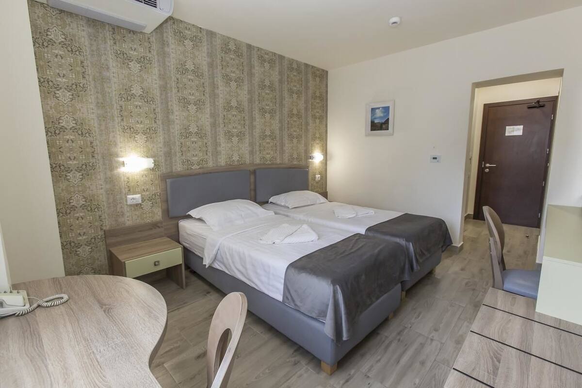 Quarto de hotel com duas camas, uma escrivaninha, uma cadeira, um guarda-roupa e uma porta.