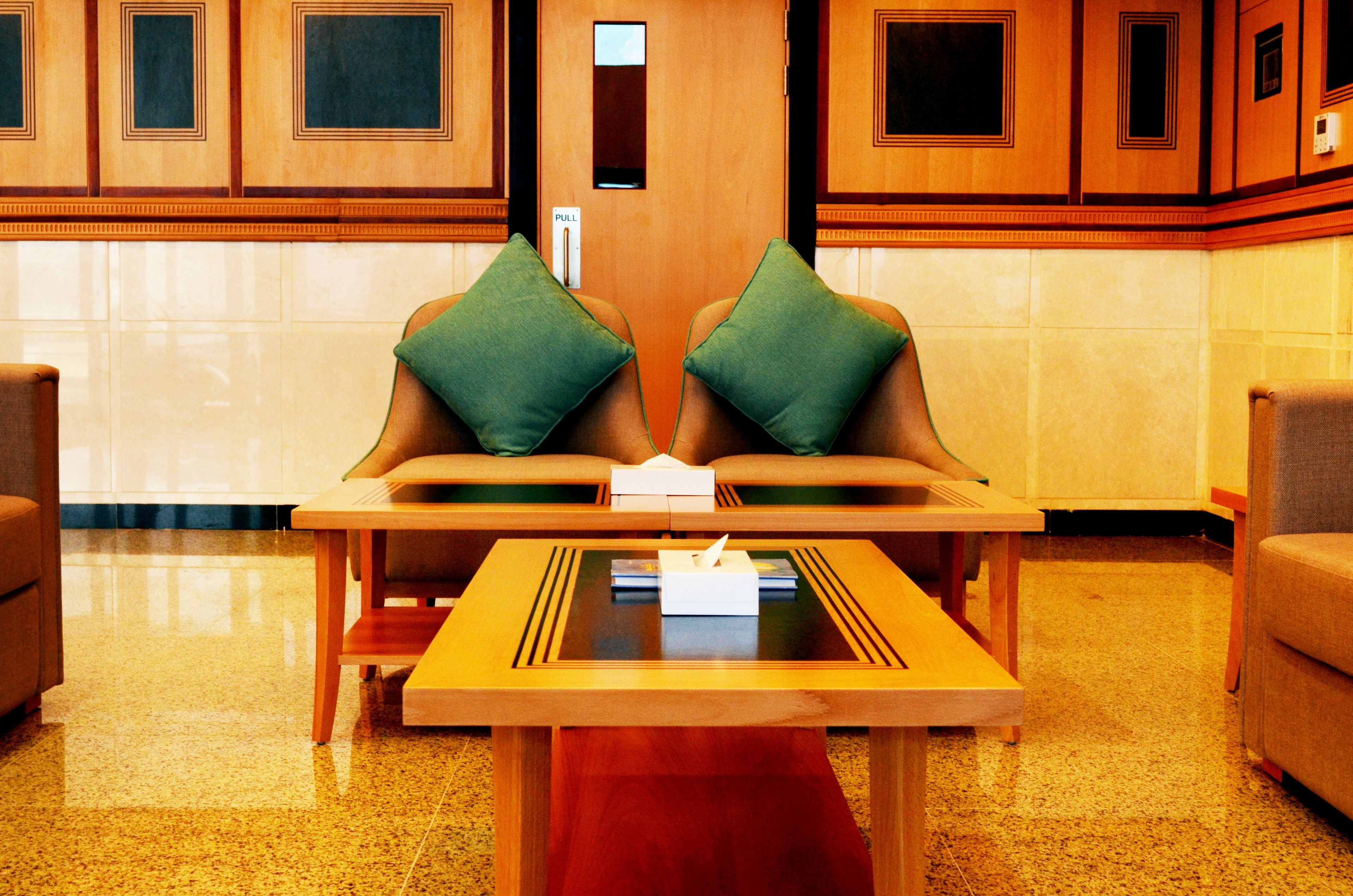Um lobby com uma mesa de recepção, duas poltronas com almofadas verdes e uma parede de madeira com a inscrição 'PULL'.