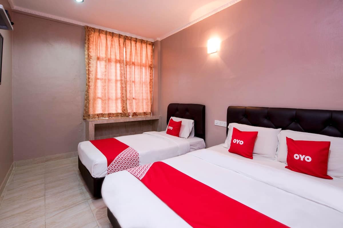 Quarto de hotel com duas camas, cada uma com roupa de cama branca e detalhes vermelhos, uma janela com cortinas florais e uma luminária fixada na parede.