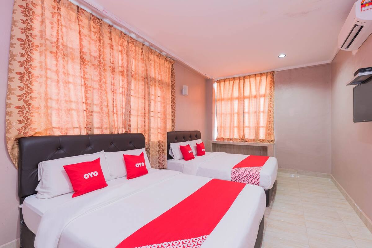 Quarto de hotel com duas camas, cada uma com colcha vermelha e branca, e uma janela grande com cortinas.