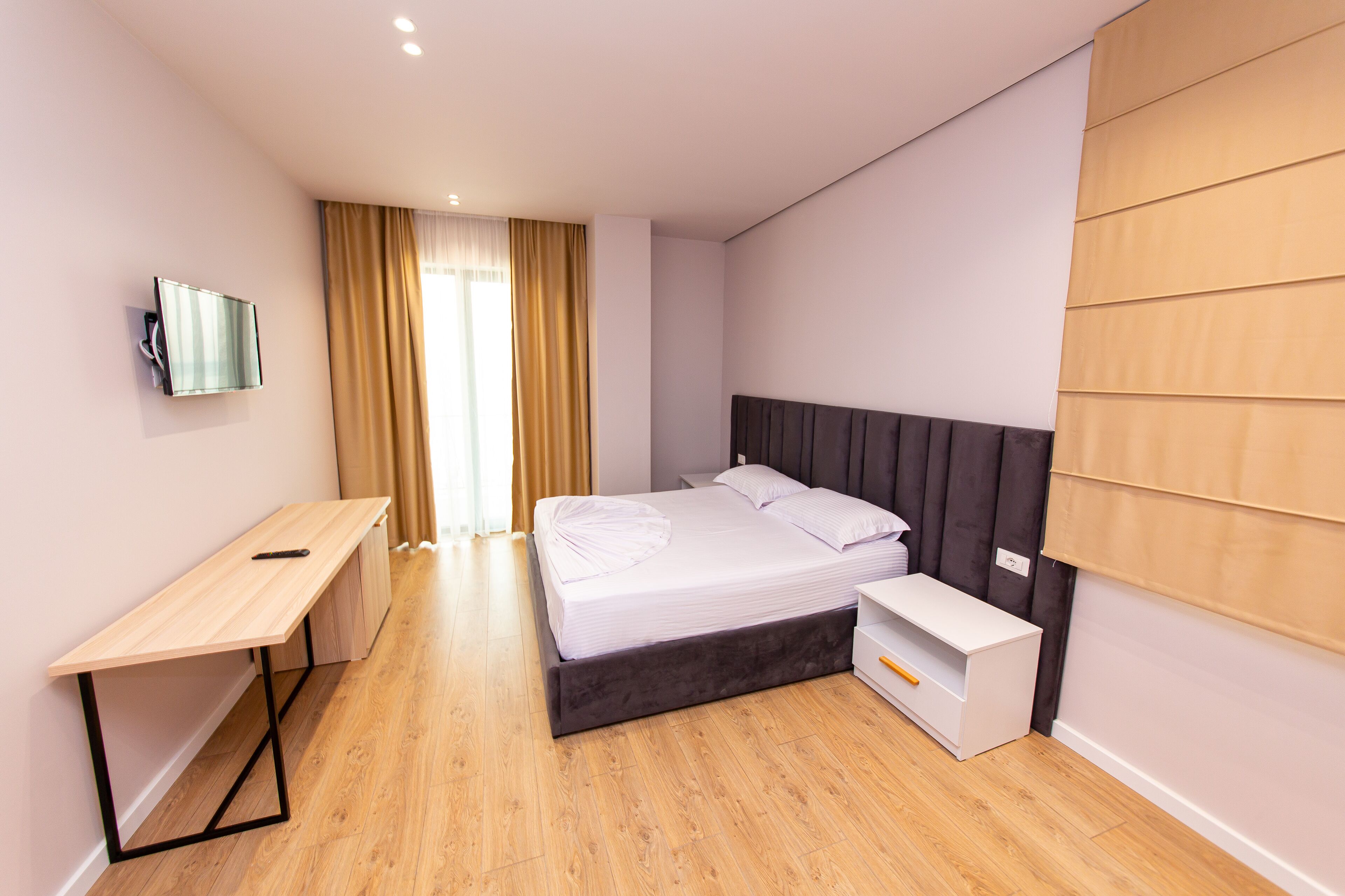 Quarto de hotel moderno com piso de madeira, cama com cabeceira escura, mesa de cabeceira, escrivaninha e televisão fixada na parede.
