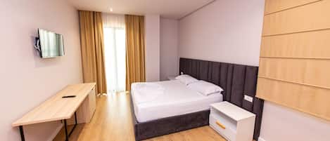 Quarto de hotel moderno com piso de madeira, cama com cabeceira escura, mesa de cabeceira, escrivaninha e televisão fixada na parede.