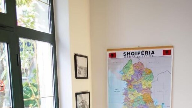 Quarto com uma janela grande, um banco verde, um mapa da Albânia e um vaso com flores.