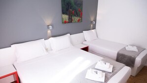 Quarto de hotel moderno com uma cama grande, uma cama menor e uma mesa de cabeceira. Há um quadro na parede e duas luminárias fixadas.