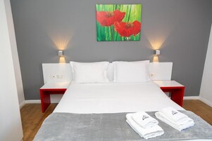 Quarto de hotel com uma cama, duas mesas de cabeceira, um banco vermelho e um quadro com flores vermelhas na parede.