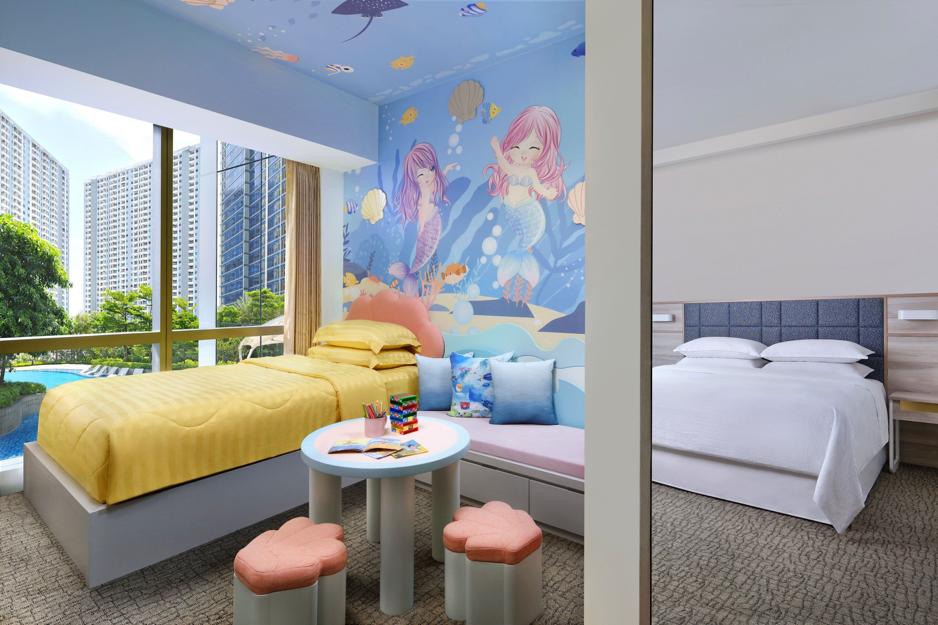 Quarto de hotel com uma cama grande, uma cama menor, uma mesa redonda com um livro e um mural de sereias na parede.