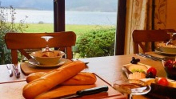 Mesa de jantar com pão, uma tigela e um prato com um item vermelho, com vista para um lago e montanhas.