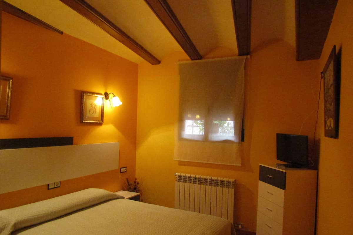 Quarto de hotel com cama, radiador, televisão sobre um móvel e janela com persianas.