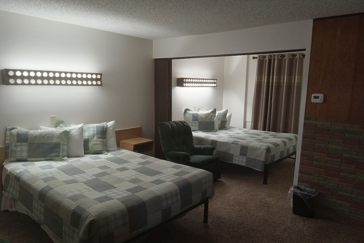 Quarto de hotel com duas camas, uma cadeira, uma mesinha e um candeeiro fixado na parede.
