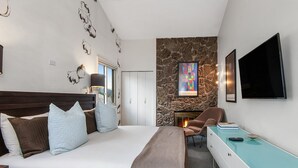 Quarto com parede de pedra, uma cama com almofadas, televisão, escrivaninha com luminária e lareira.