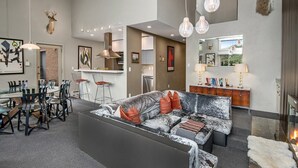 Uma sala de estar moderna com um amplo sofá de canto, uma área de jantar com mesa de vidro e cadeiras de metal, e uma cozinha ao fundo.