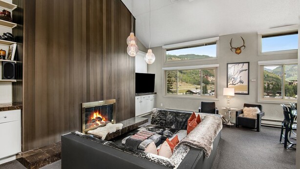 Uma sala de estar moderna com lareira, sofá, TV e janelas amplas com vista para o exterior.