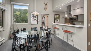 Uma cozinha moderna com uma ilha grande, uma área de refeições com mesa de vidro e cadeiras pretas, e vista para uma floresta pela janela.