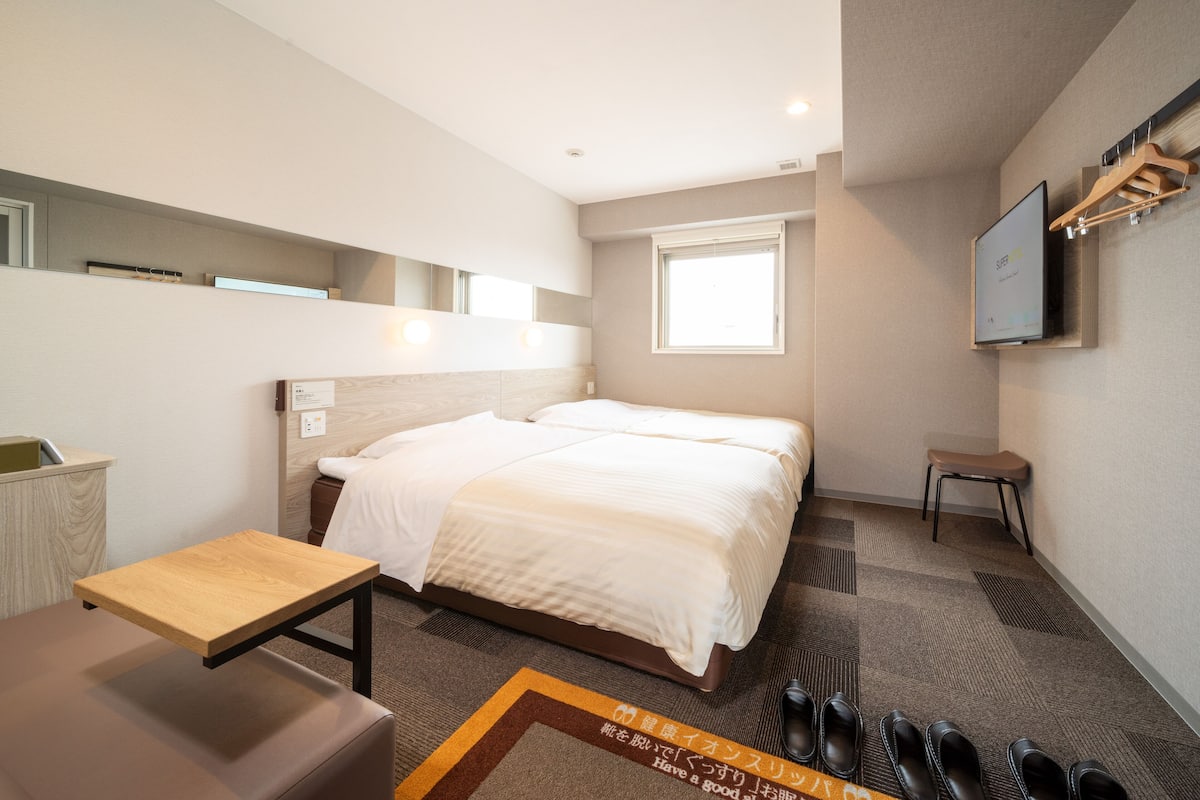Quarto de hotel com cama, escrivaninha, cadeira, televisão fixada na parede e sapateira.