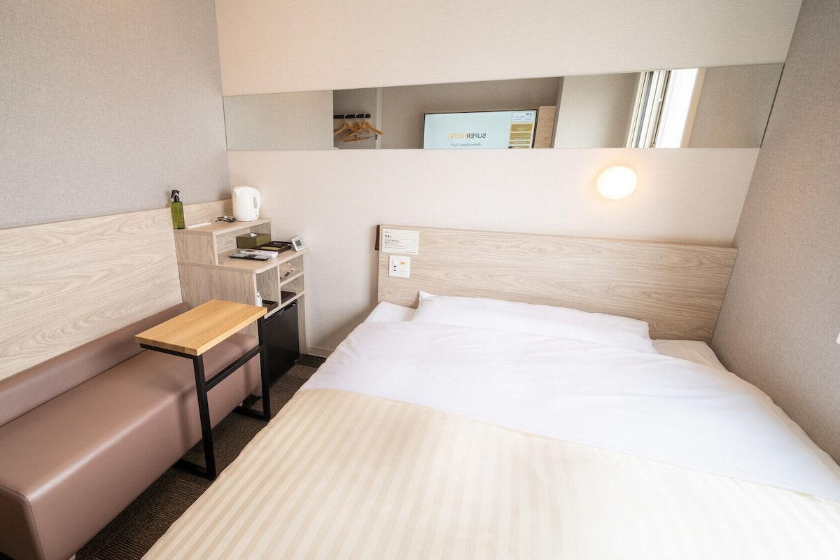 Quarto de hotel com cama, escrivaninha, um banco, prateleira pequena com artigos de higiene pessoal e um espelho.