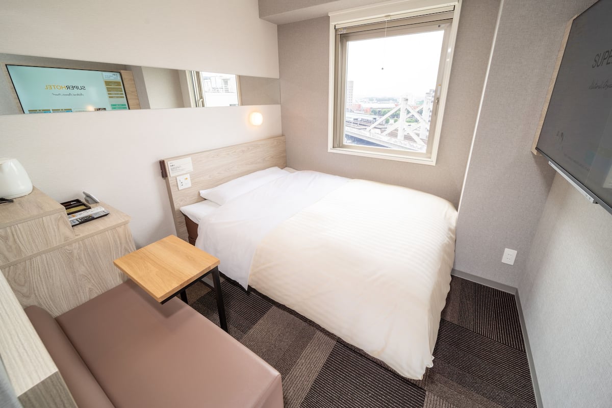 Quarto de hotel com cama, escrivaninha, cadeira, janela com vista para a cidade e televisão presa à parede.