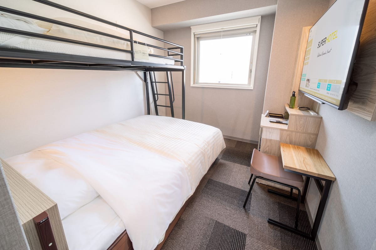 Quarto de hotel compacto com beliche, escrivaninha, cadeira, janela pequena e televisão fixada na parede.