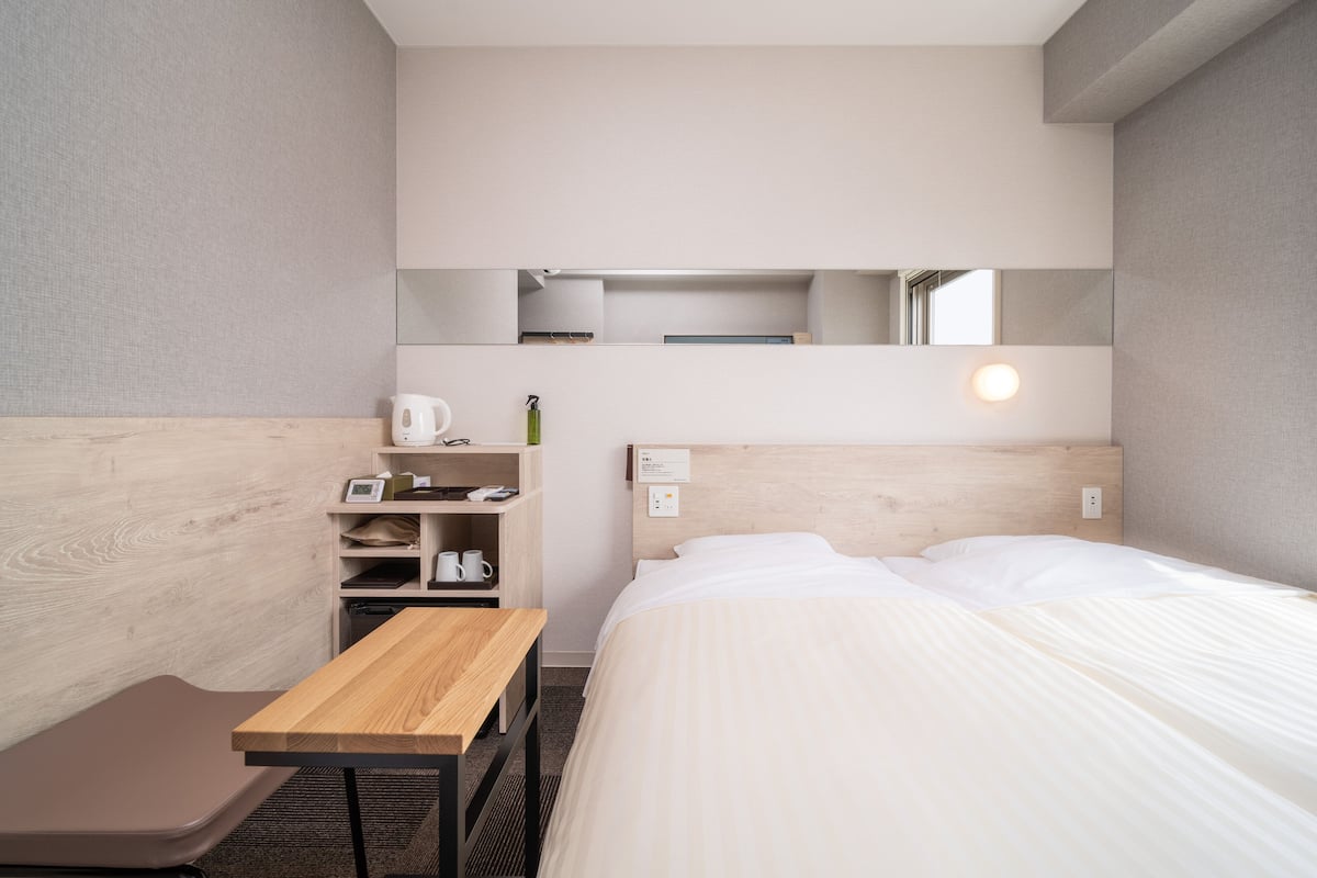 Quarto de hotel com cama, escrivaninha, cadeira, mesinha, espelho e prateleira com produtos de higiene pessoal.