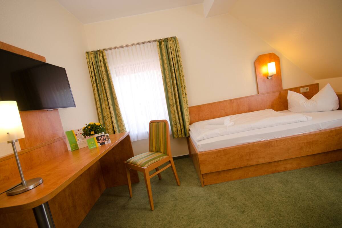 Quarto de hotel com cama, escrivaninha com luminária, cadeira, televisão e janela com cortinas.