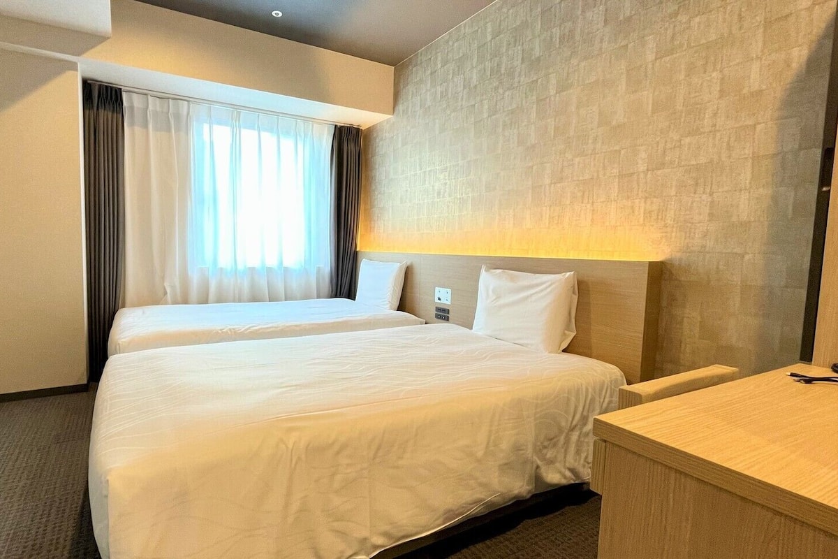 Quarto de hotel com duas camas, uma televisão de tela plana, uma escrivaninha e uma janela com cortinas.