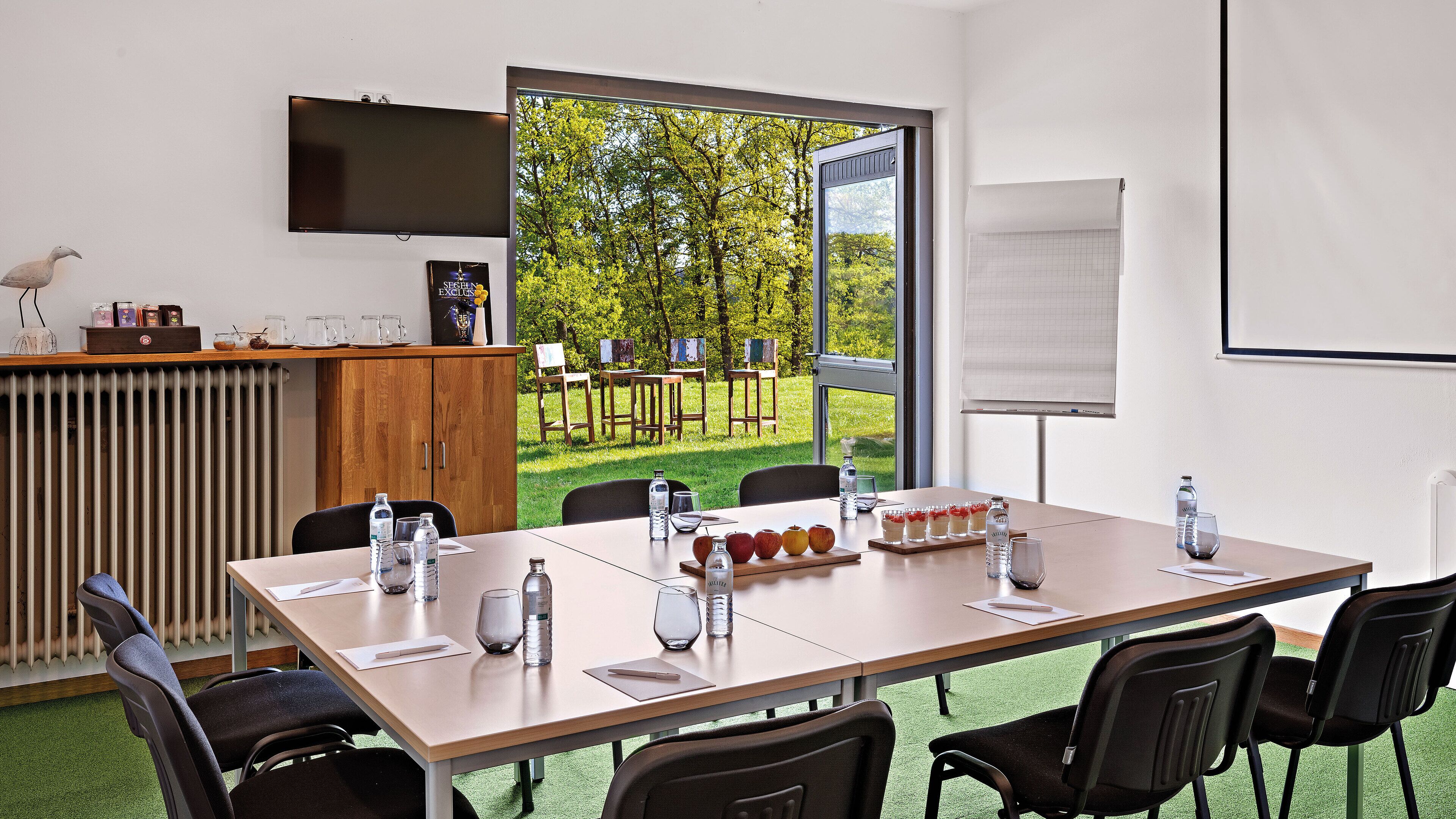Uma sala de conferências com uma mesa grande, cadeiras, uma tela de projetor e vista para um jardim pelas janelas.