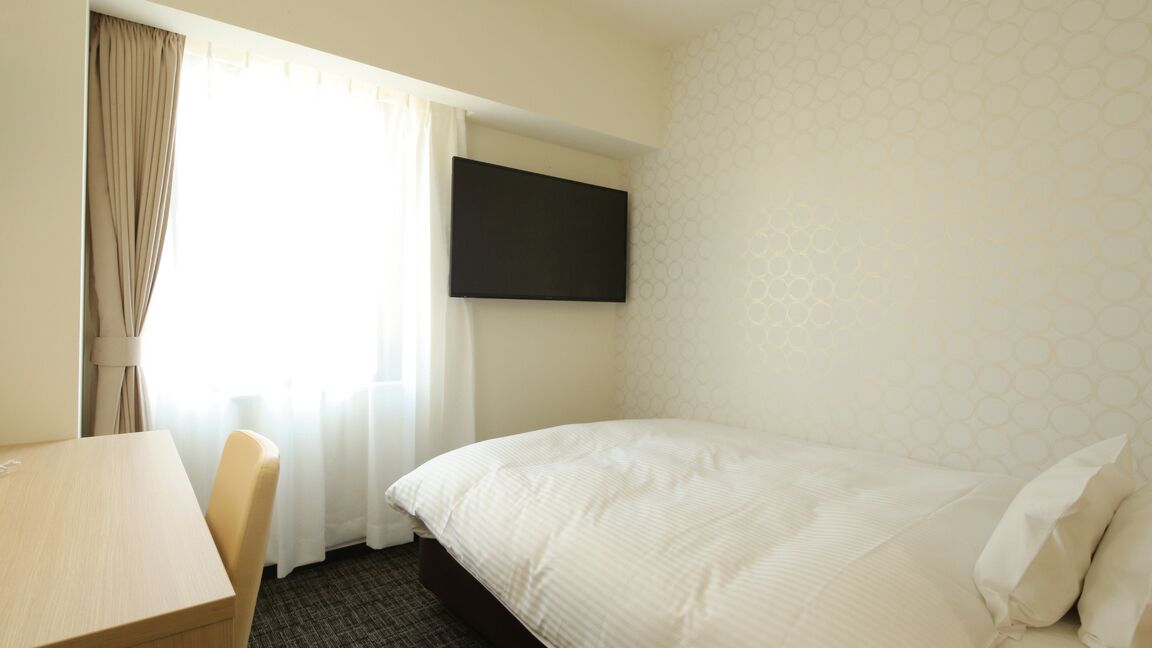Quarto de hotel com uma cama, televisão fixada na parede, janela com cortinas e uma pequena mesa.