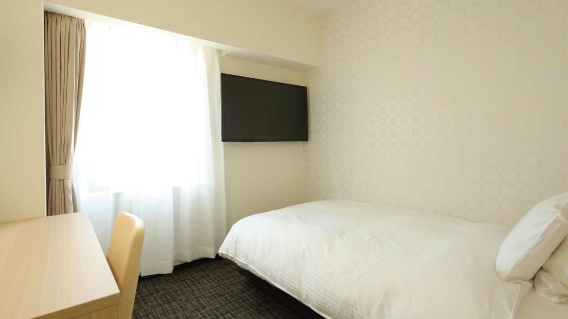 Quarto de hotel com cama, televisão na parede, escrivaninha e uma cadeira.