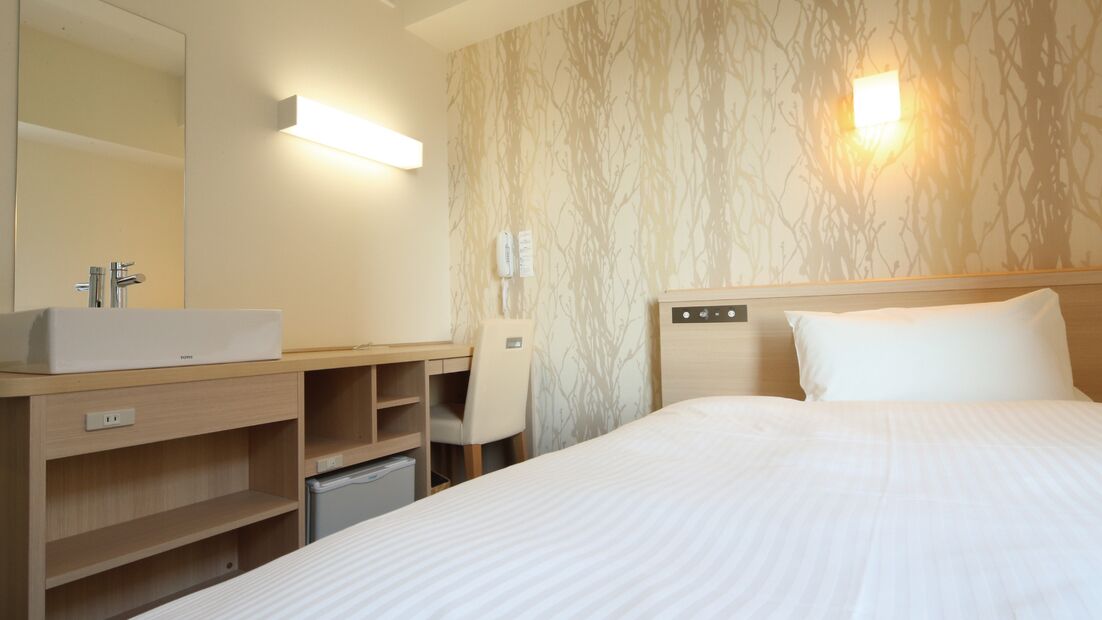 Quarto de hotel com cama, mesa de cabeceira, espelho, escrivaninha com cadeira e luminária na parede.