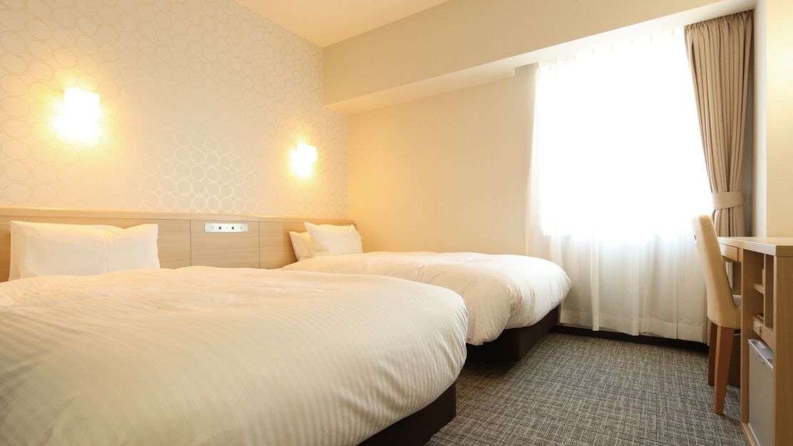 Quarto de hotel com duas camas, uma escrivaninha e uma janela com cortinas.