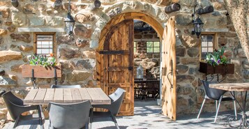 Paredes de pedra externas com uma porta de madeira, uma mesa com cadeiras e lanternas penduradas.