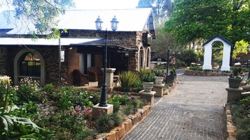 Um edifício de pedra com telhado ladrilhado, um jardim com diversas plantas e flores, e um caminho de pedras que leva a um arco branco.