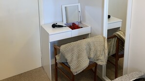 Quarto pequeno e arrumado, com uma toucador branca, espelho e uma cadeira com manta.