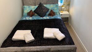 Cama bem arrumada com cabeceira cinza, roupa de cama floral azul e duas toalhas brancas.