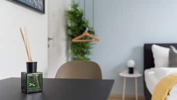 Quarto de hotel moderno com mesa preta, planta pendente, mesinha redonda com luminária branca e cama com roupa de cama branca.