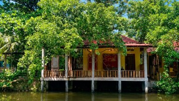 Uma casa com telhado vermelho, sustentada por estacas sobre a água, rodeada por vegetação exuberante.