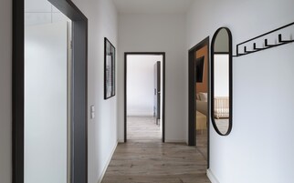 Um corredor com piso de madeira, uma porta que dá para um quarto com cama e berço, e um espelho fixado na parede.