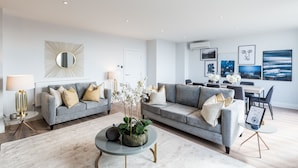Uma sala de estar moderna com dois sofás cinzentos, uma mesa de centro com objetos decorativos e uma área de jantar com cadeiras e mesa.