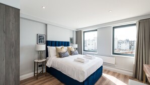 Quarto de hotel moderno com uma cama grande, uma mesinha de cabeceira e vista para a cidade através de uma janela ampla.