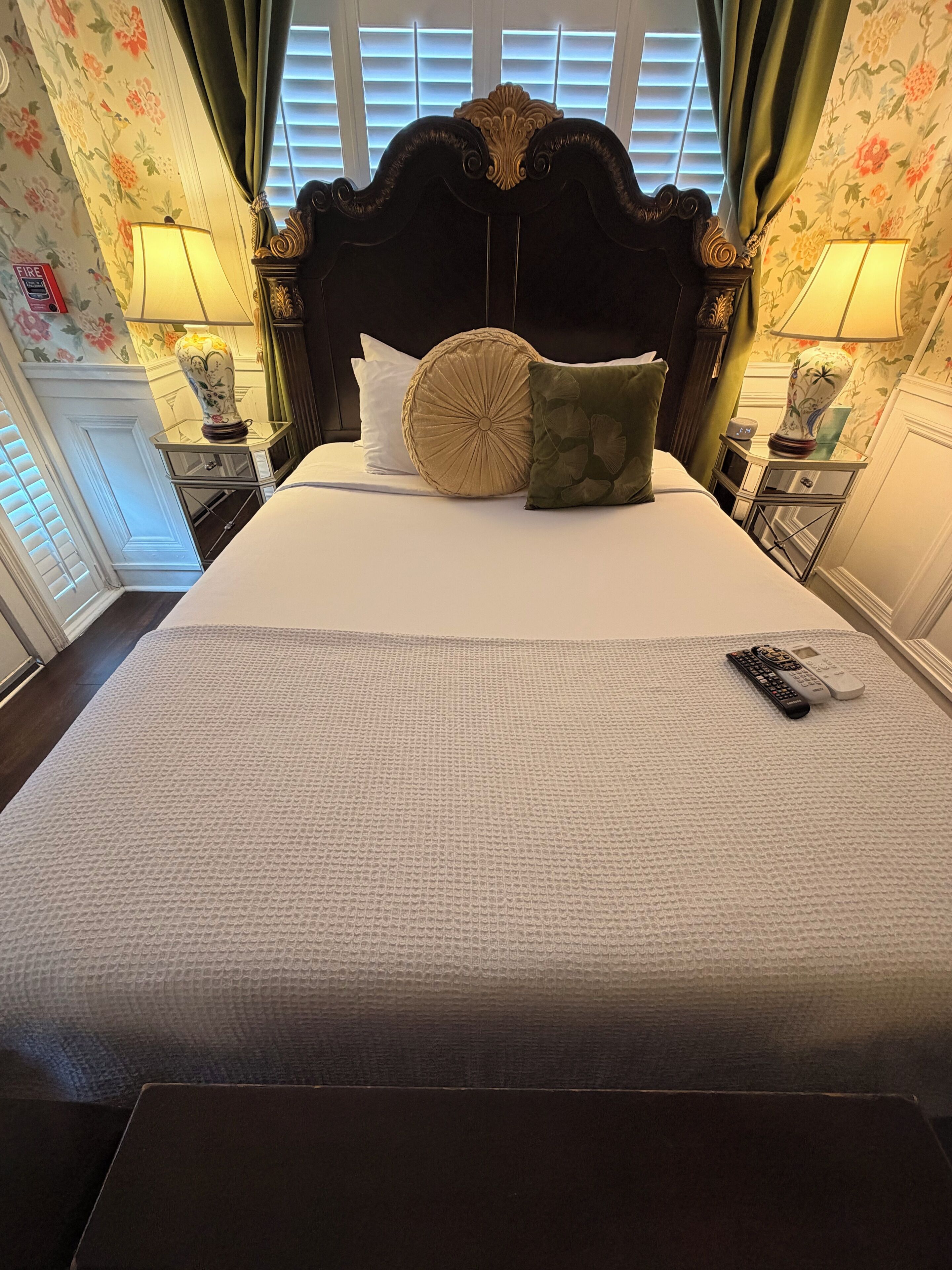 Cama bem arrumada com cabeceira, duas luminárias de apoio, um controle remoto e papel de parede floral.