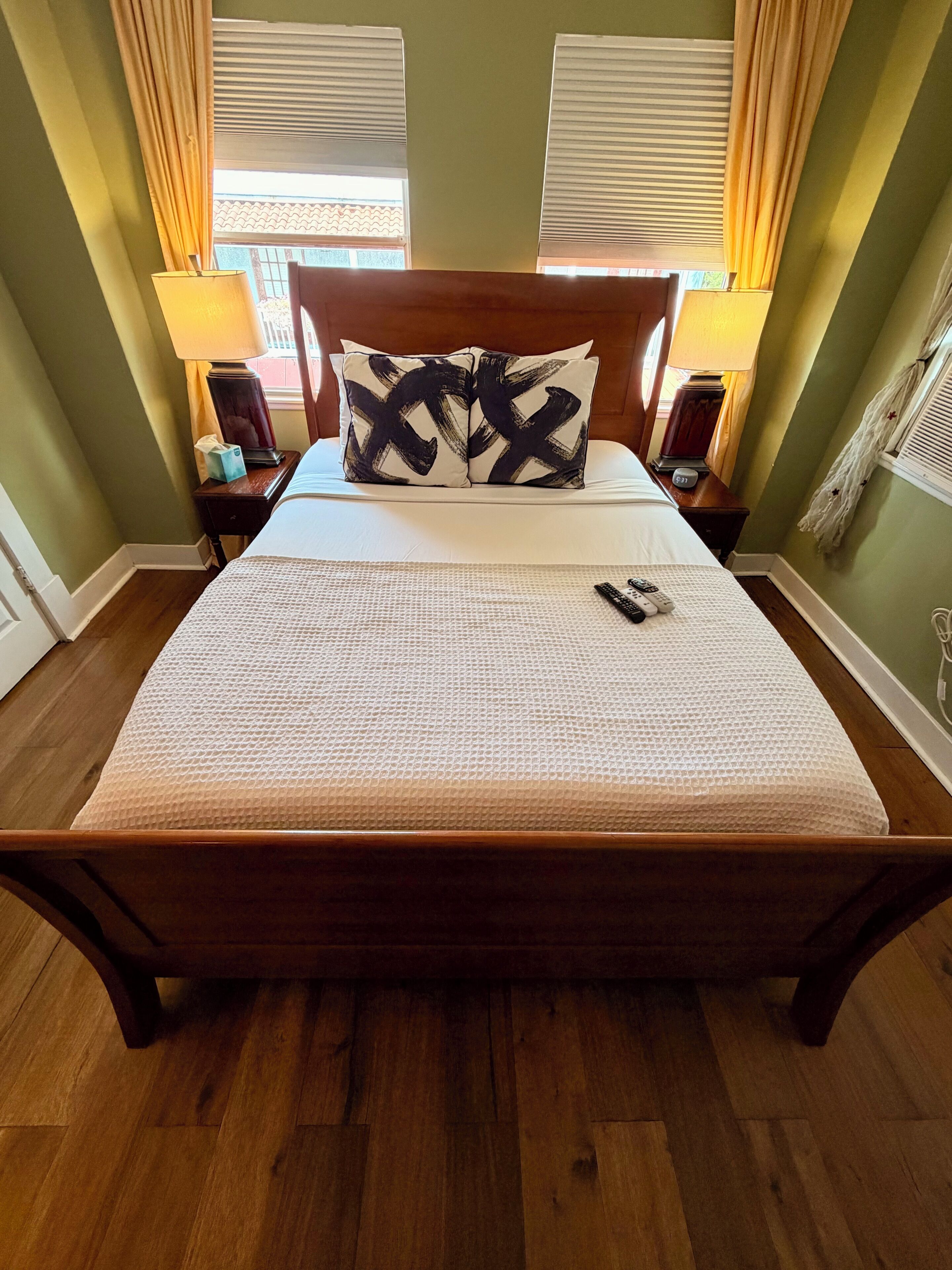 Uma cama com colchão branco texturizado, duas almofadas com padrão preto e branco, cabeceira de madeira, duas mesas de cabeceira com abajures e piso de madeira.