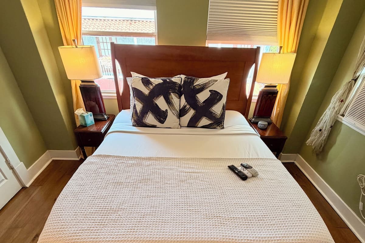 Uma cama com colchão branco texturizado, duas almofadas com padrão preto e branco, cabeceira de madeira, duas mesas de cabeceira com abajures e piso de madeira.