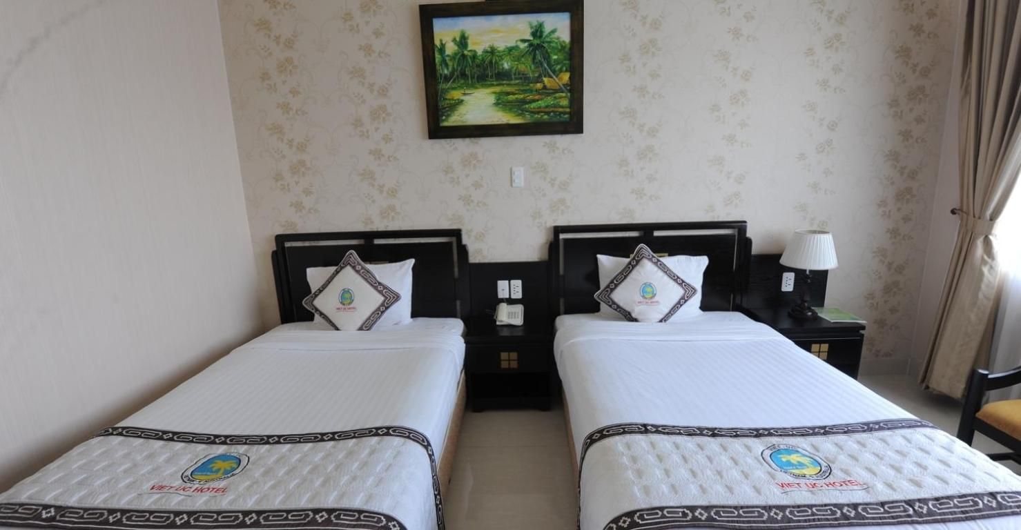 Quarto de hotel com duas camas, mesa de cabeceira com luminária, pintura na parede e janela com cortinas.