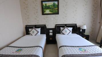 Quarto de hotel com duas camas, mesa de cabeceira com luminária, pintura na parede e janela com cortinas.