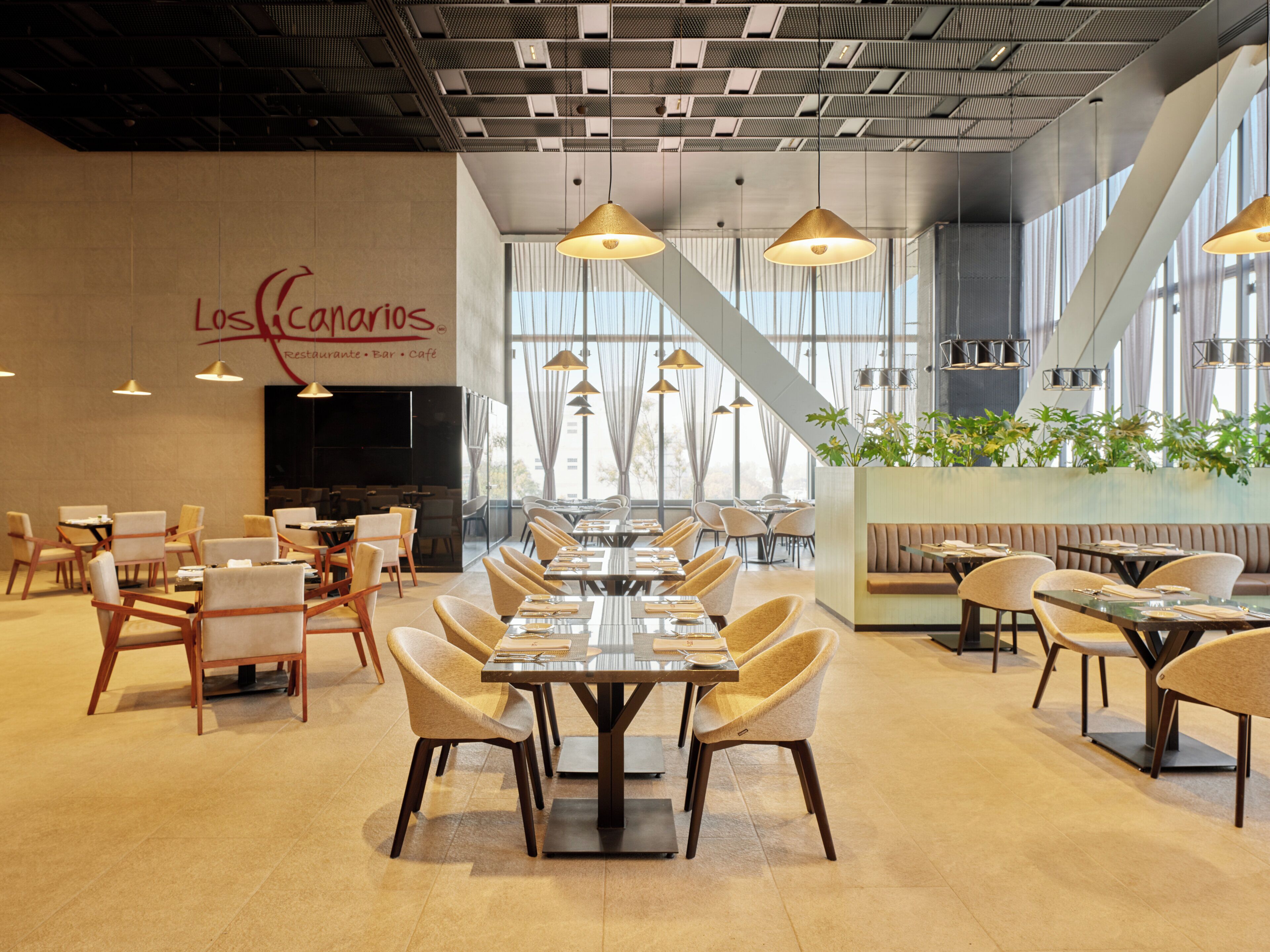 Uma sala de jantar moderna com várias mesas e cadeiras, uma grande janela com cortinas e uma placa indicando 'Los Canarios Restaurante Bar Café'.