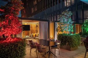 Uma área de refeições ao ar livre com mesa e cadeiras, rodeada por vegetação e um edifício moderno ao fundo.