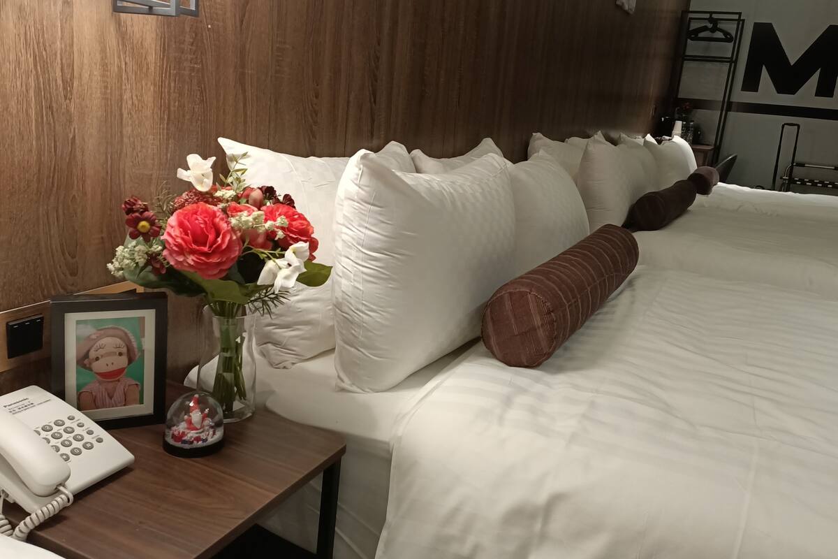 Quarto de hotel com cabeceira de madeira, roupa de cama branca, mesa de cabeceira com telefone, moldura de quadro e um vaso com flores.
