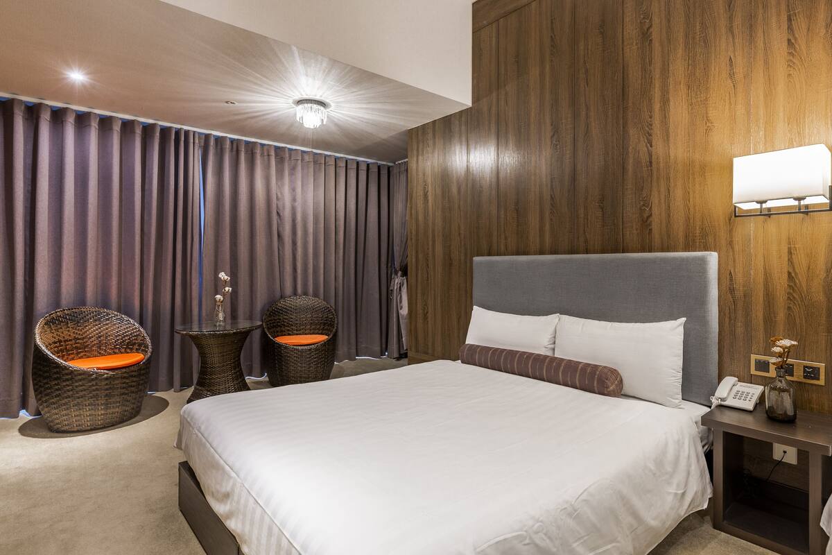 Quarto de hotel com uma cama grande, duas cadeiras de vime com almofadas laranja, uma mesinha de cabeceira com abajur e uma parede de destaque em madeira.