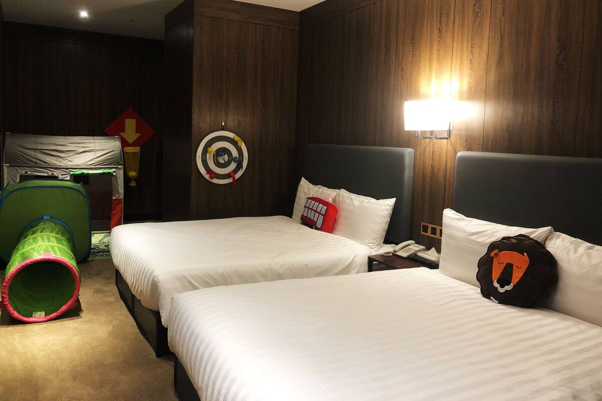 Quarto de hotel com duas camas, um alvo na parede e um brinquedo de túnel verde.