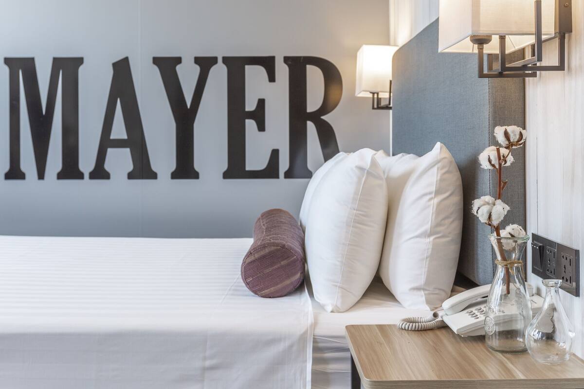 Quarto de hotel com uma cama grande, uma mesa de cabeceira com abajur, um vaso com flores e uma parede com a palavra "MAYER" em letras grandes.