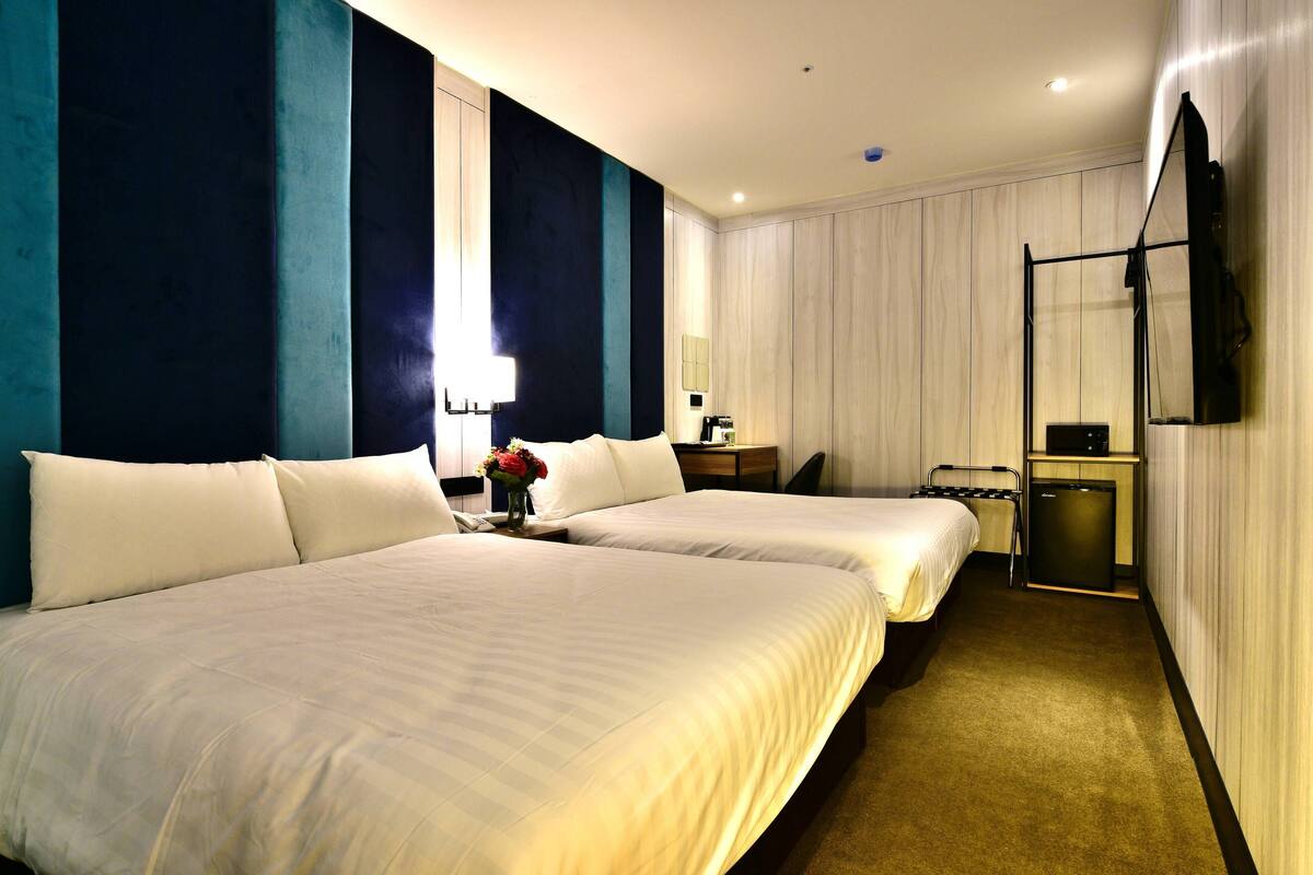 Quarto de hotel com duas camas, uma escrivaninha, televisão presa à parede e uma pequena cozinha.